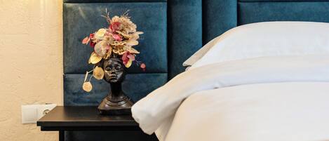 Cama bem arrumada com lençóis brancos, cabeceira estofada em azul escuro e mesa de cabeceira com um vaso de flores.