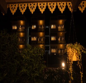 Um edifício de vários andares com janelas e varandas iluminadas, cercado por vegetação densa.