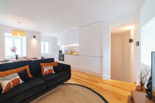 Um apartamento moderno com sofá escuro, cozinha com armários brancos e piso de madeira.