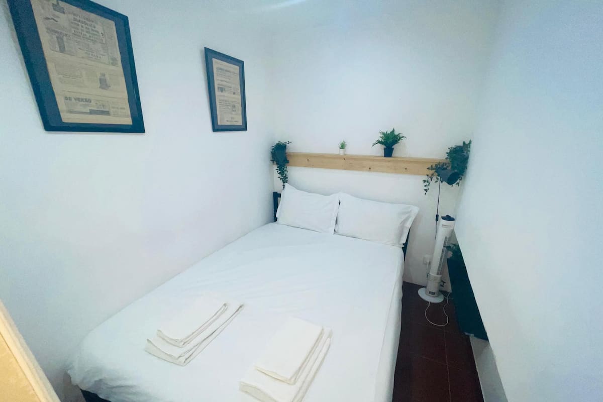 Uma cama pequena e bem arrumada, com lençóis e toalhas brancos, cabeceira de madeira e uma pequena prateleira com uma planta.