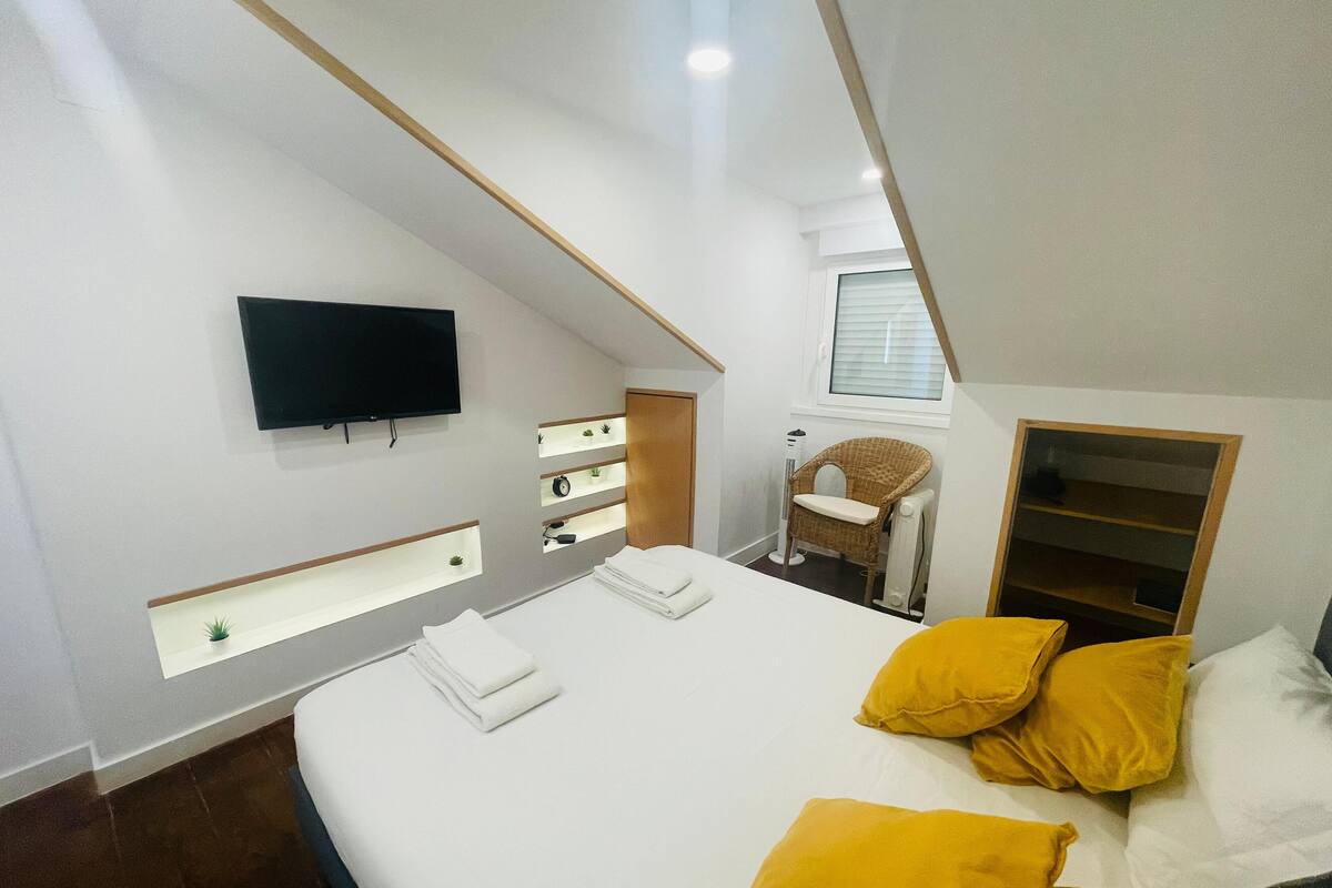 Quarto de hotel moderno com cama, televisão, prateleiras embutidas, uma cadeira e janela.