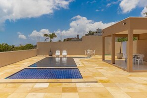 Uma área de piscina no terraço com uma parte em azulejos azuis, cadeiras brancas e um espaço coberto para sentar.