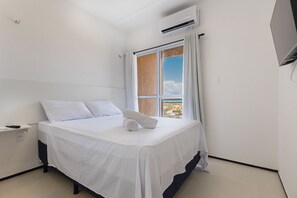 Cama bem arrumada com lençóis brancos, janela com cortinas e ar condicionado na parede.