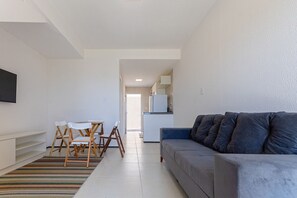 Uma sala de estar moderna com um sofá cinza, cadeiras de madeira, um tapete listrado e uma cozinha ao fundo.