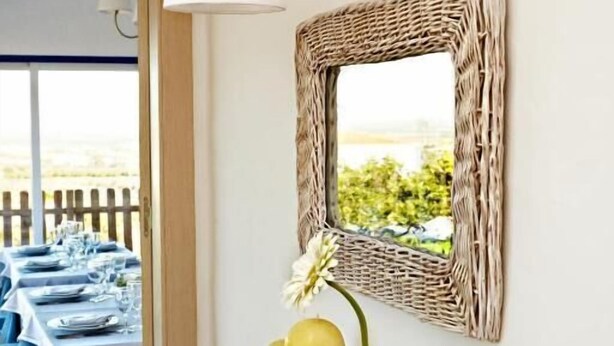 Quarto com área de jantar visível através de uma porta aberta, luminária de parede, espelho com moldura de vime e um pequeno estante com objetos decorativos.