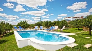 Uma piscina retangular com borda curva, cercada por gramado e árvores. Há cadeiras de descanso ao longo da borda da piscina.
