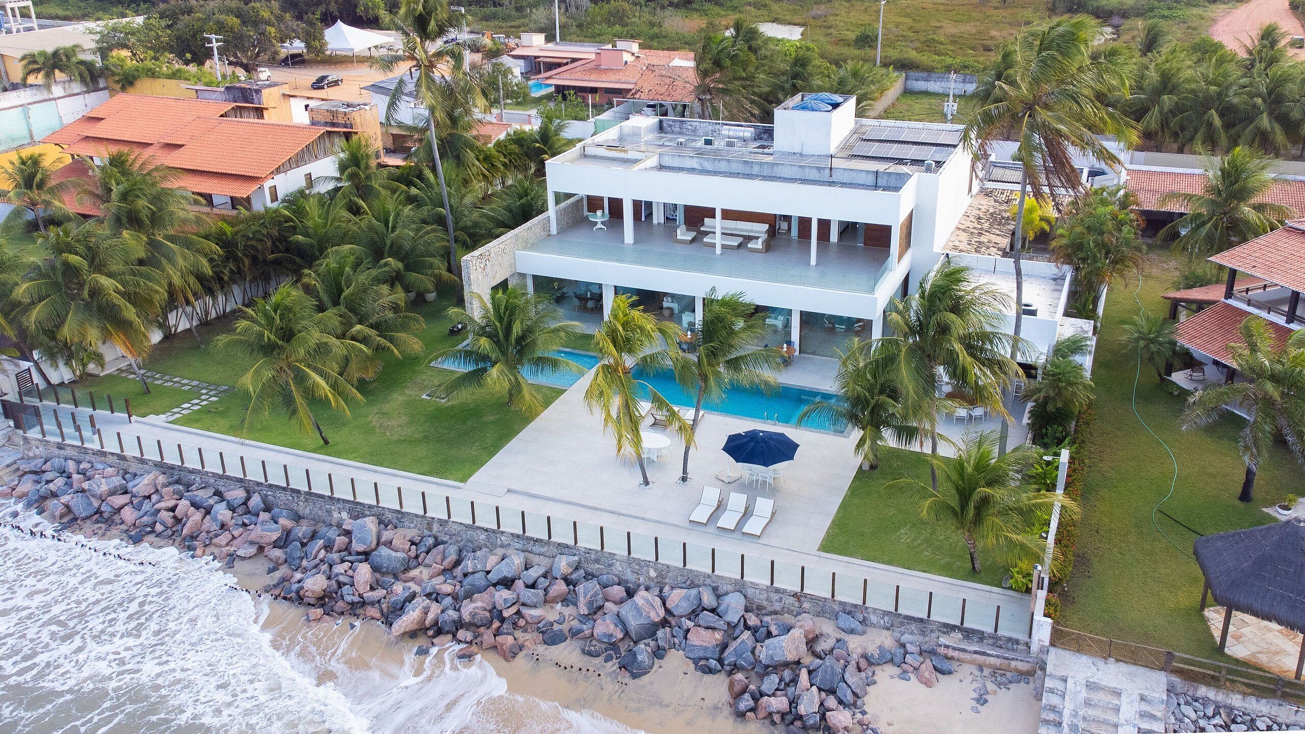 Vista aérea de uma propriedade moderna à beira-mar, com piscina, palmeiras e um trecho de costa rochosa.