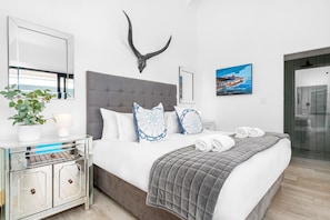 Quarto de hotel moderno com uma cama grande, cabeceira cinza, almofadas decorativas, mesa de cabeceira com abajur, espelho e uma grande obra de arte na parede.