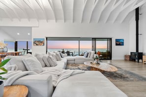 Uma sala de estar moderna com um amplo sofá de canto, uma mesa de centro e vista para o oceano ao pôr do sol.