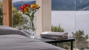 Uma cama com vista para um lago e montanhas, um vaso com flores e uma pilha de livros numa mesa de cabeceira.