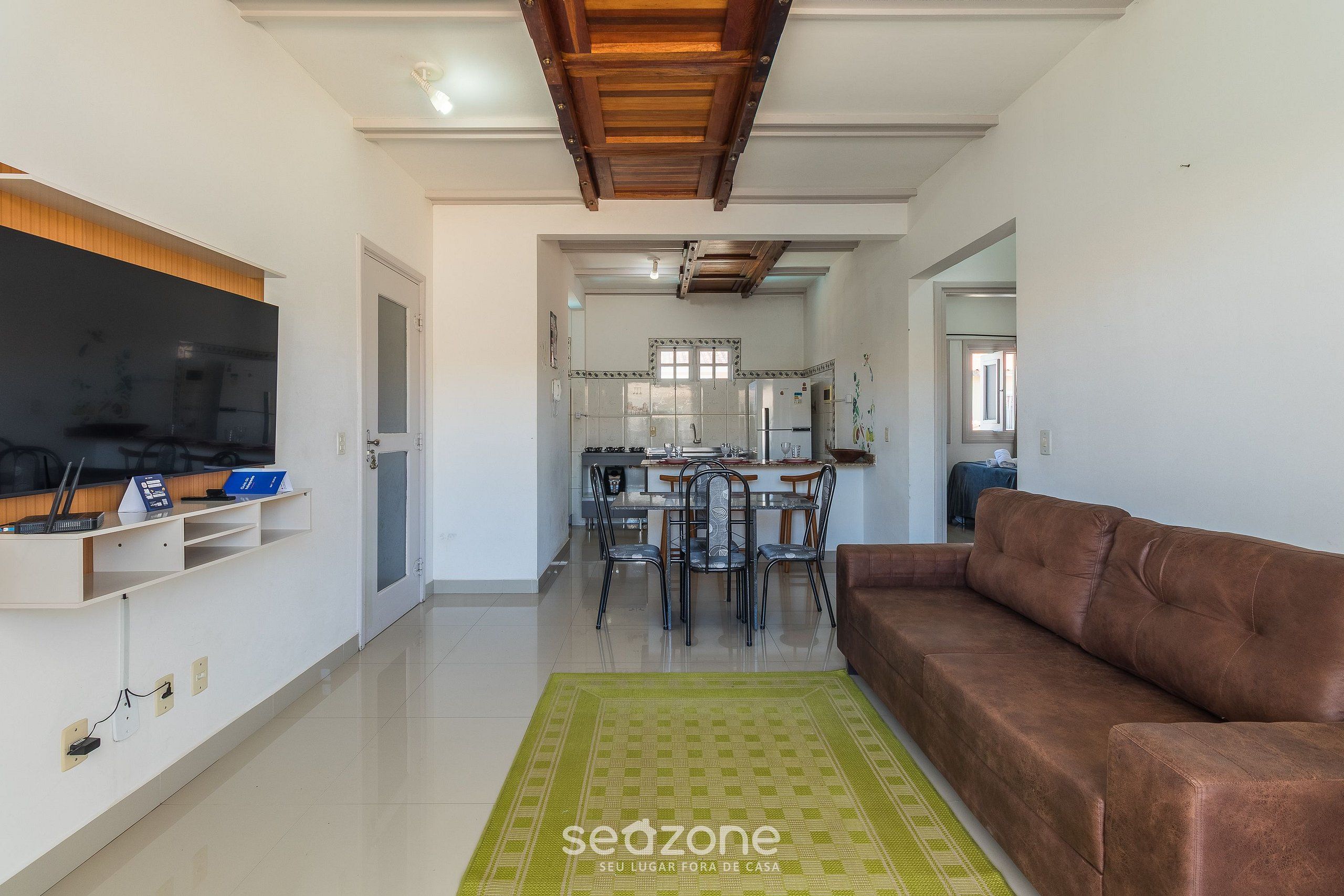 Uma sala de estar moderna com um sofá marrom, um tapete verde, uma TV fixada na parede e uma área de jantar com cadeiras e uma mesa.