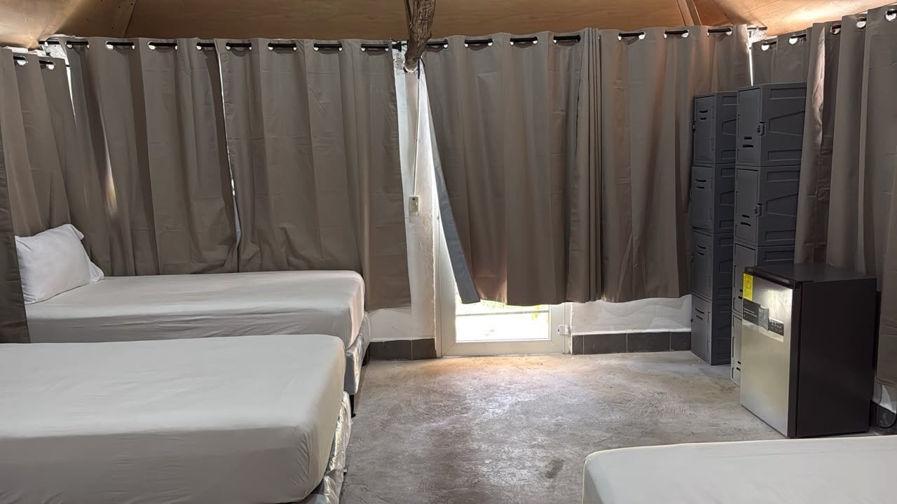 Quarto com duas camas, um pequeno espaço de cozinha e uma janela com cortinas.