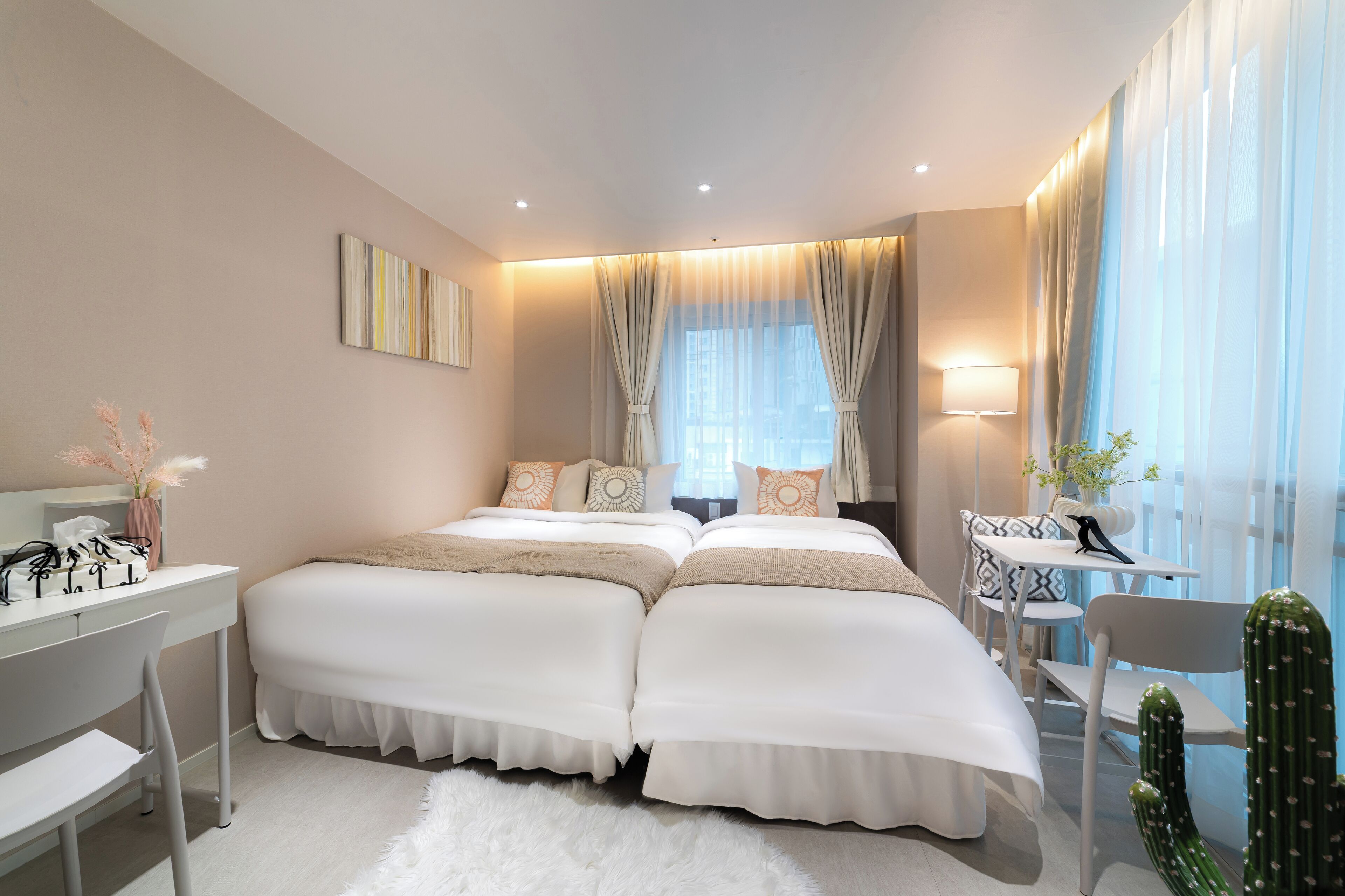 Quarto moderno com uma cama grande, uma escrivaninha e uma cadeira. Há uma janela com cortinas leves e um abajur na parede.