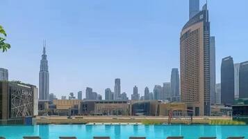 Vista da piscina com espreguiçadeiras, um edifício moderno com fachada singular e um horizonte urbano ao fundo.
