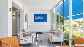 Uma área de espera moderna com cadeiras brancas, uma mesa preta e uma janela grande com toldo azul. Uma citação de Helen Keller está exposta na parede.
