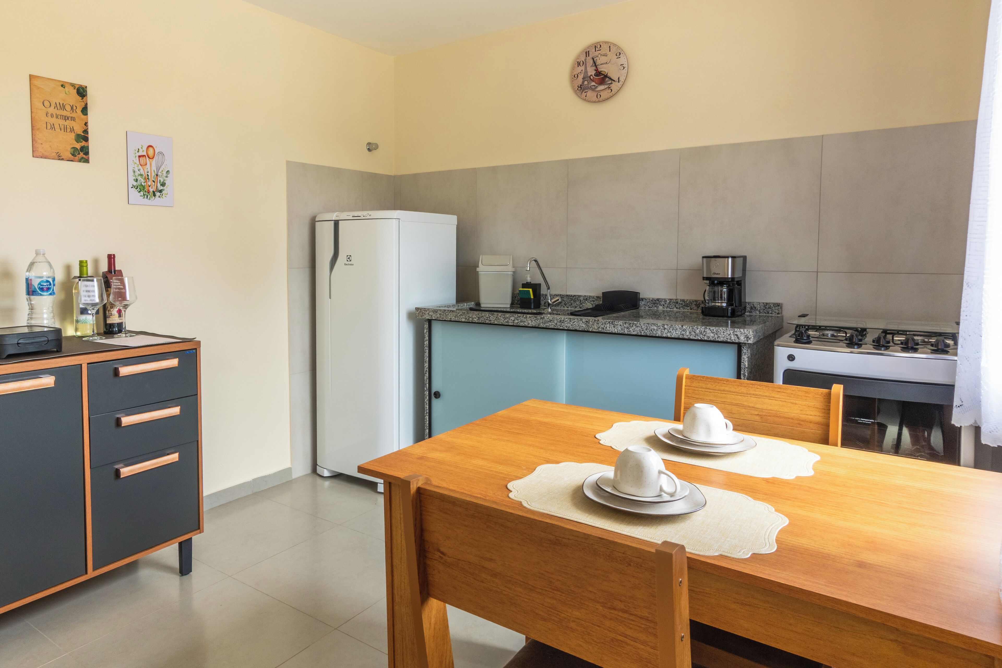 Uma cozinha equipada com geladeira, fogão e bancada. Há uma mesa de jantar com cadeiras e uma cafeteira.