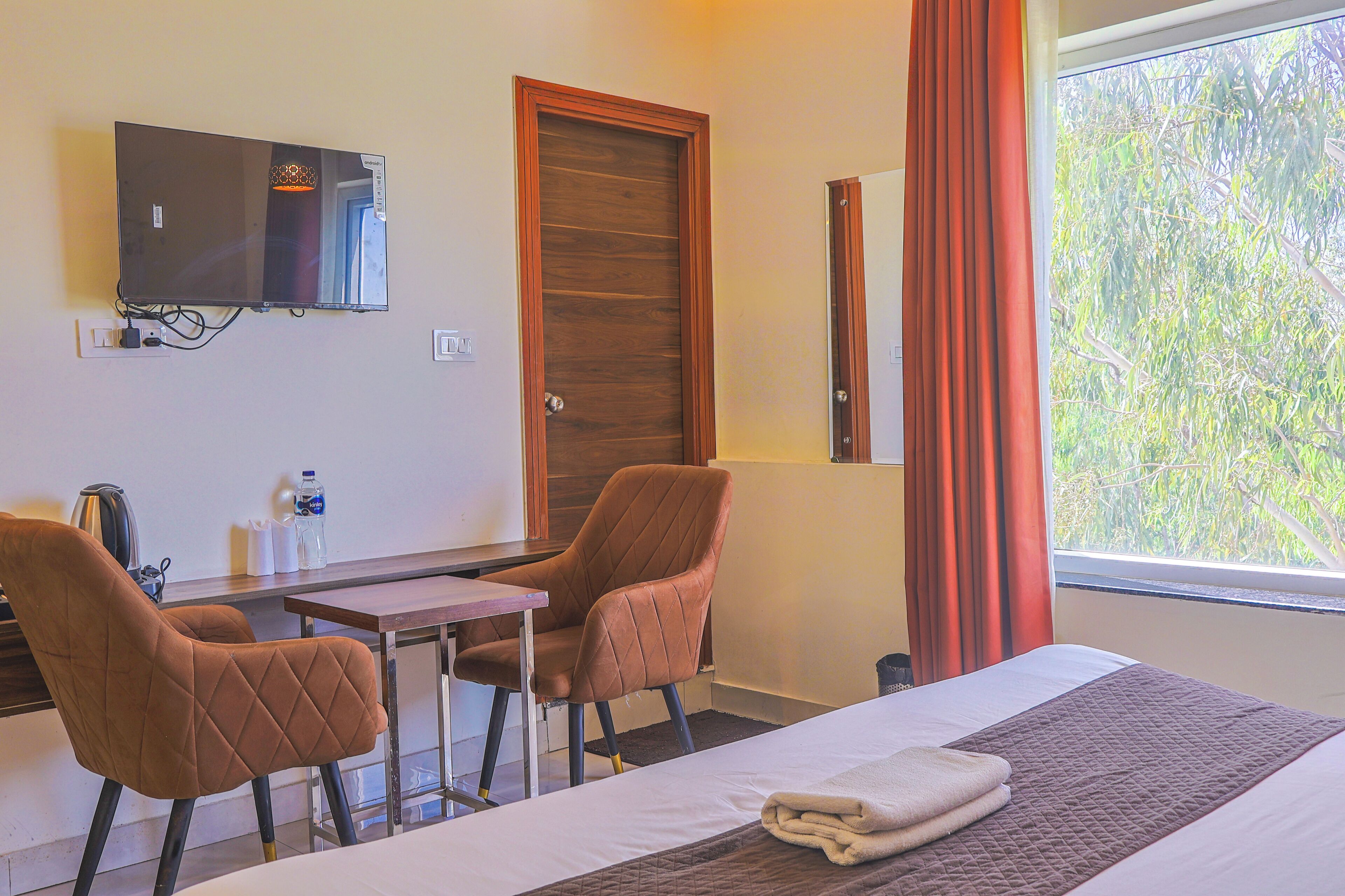 Quarto de hotel com cama, duas cadeiras, escrivaninha, televisão e janela com cortinas.
