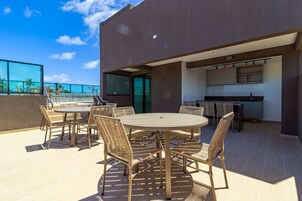 Um terraço no telhado com uma mesa redonda e cadeiras de vime, uma área de cozinha com armários, e vista para o mar.