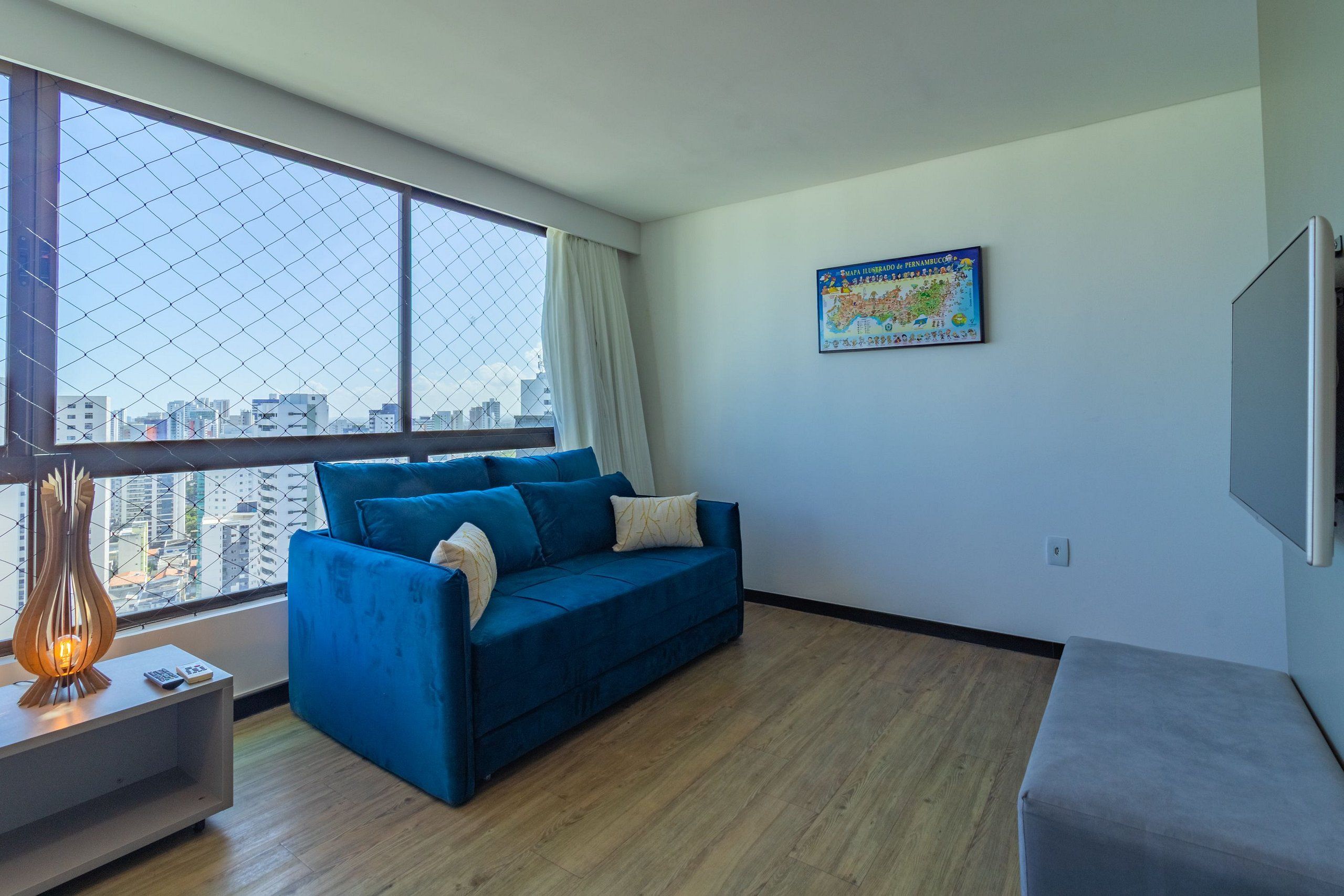Uma sala de estar moderna com um sofá azul, uma pequena mesa lateral com uma luminária, um mapa na parede e uma grande janela com vista para a cidade.