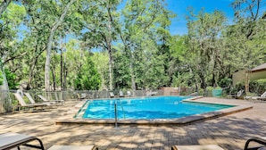 Piscina retangular com deck de tijolos, rodeada por cadeiras de descanso e árvores.