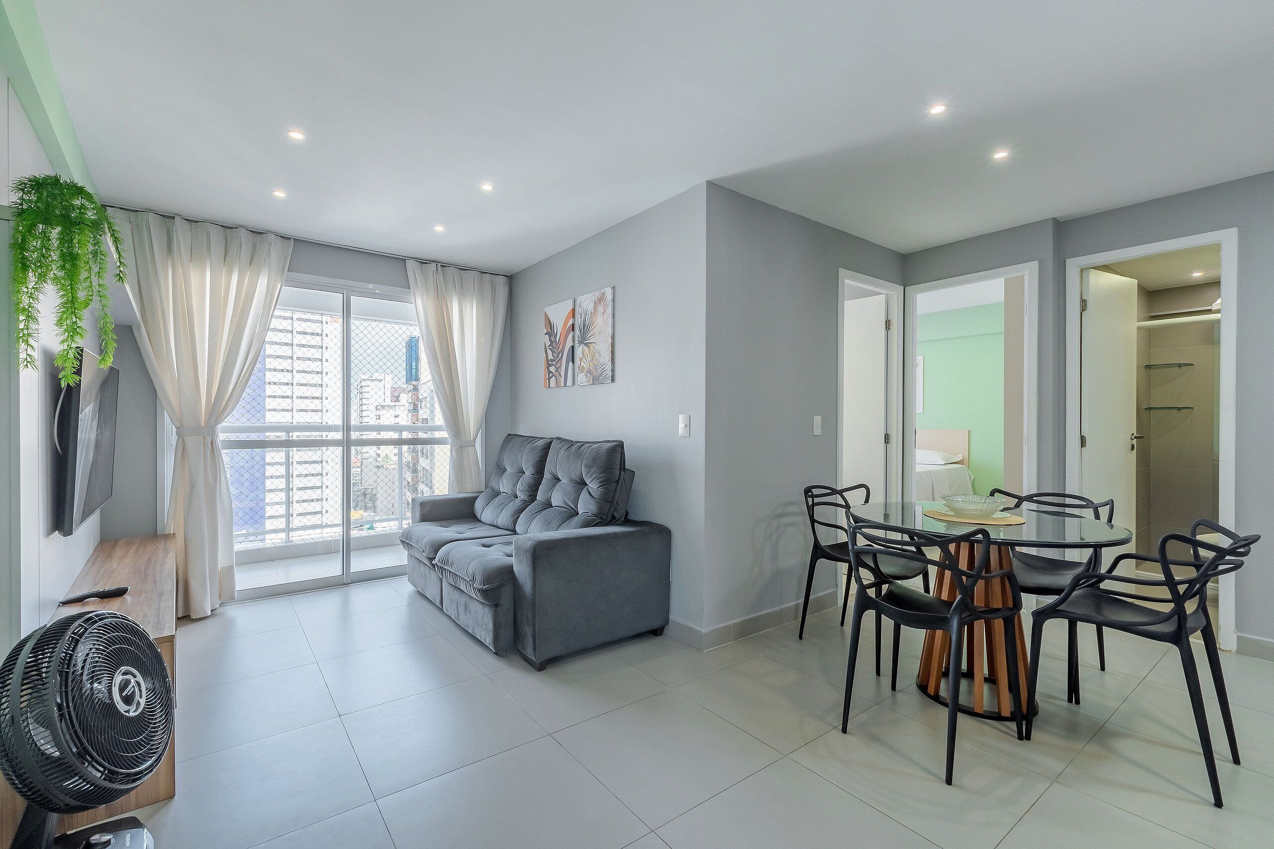 Uma sala de estar moderna com um sofá cinzento, uma mesa de jantar com cadeiras, um ventilador e uma janela ampla com cortinas.