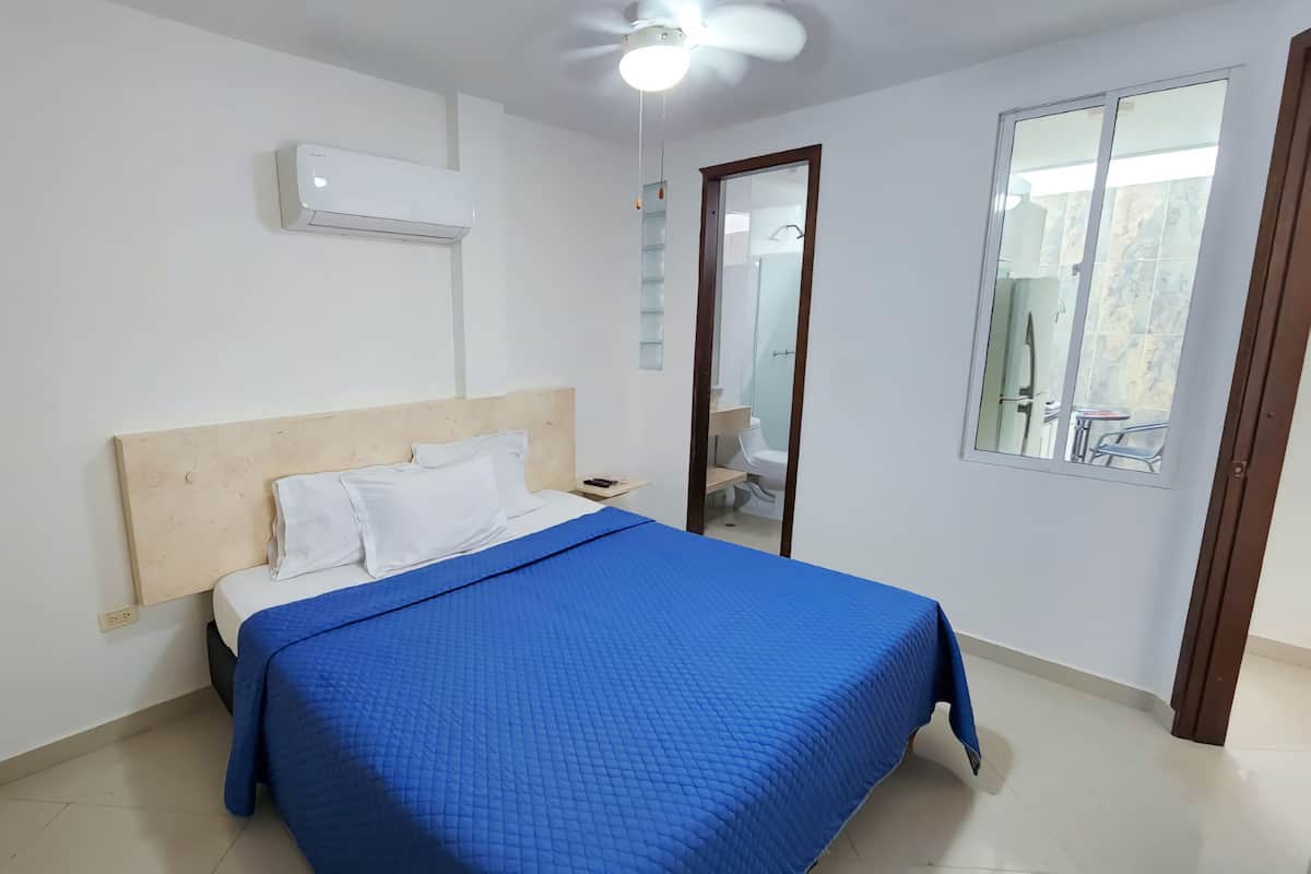 Quarto com cama, ar condicionado, ventoinha no teto, janela e porta que dá para um banheiro.