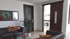 Uma sala de estar com sofá, mesa de centro de vidro com flores, planta em vaso e vista para a cozinha.