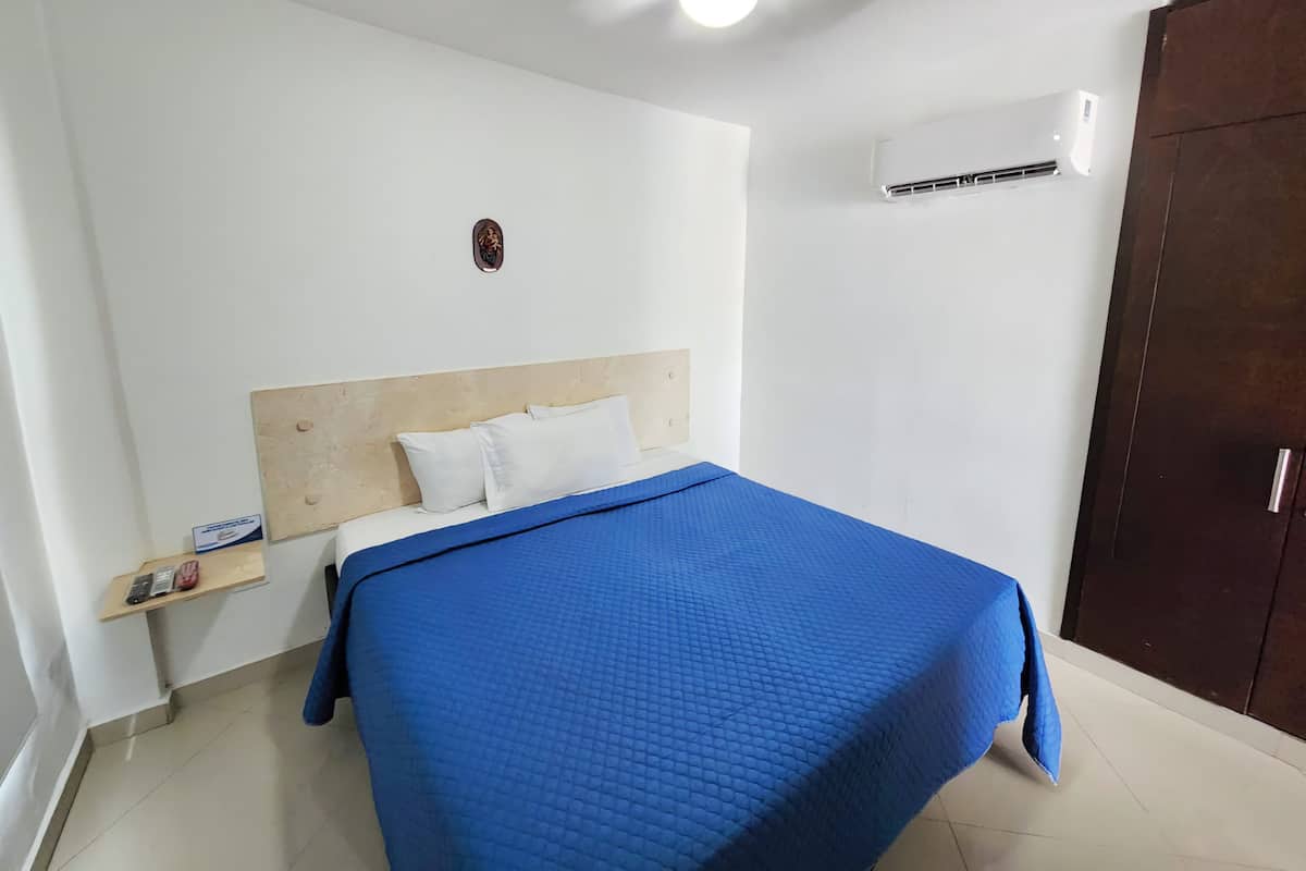 Quarto com cama, cabeceira de madeira, mesa de cabeceira, prateleira com objetos, guarda-roupa e aparelho de ar condicionado.