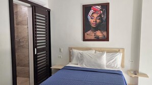 Um quarto com cama, cabeceira, mesa de cabeceira, mesa lateral, um quadro pendurado na parede e uma porta com persiana.
