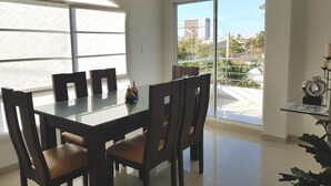 Uma área de refeições com mesa e cadeiras de vidro, janelas amplas com vista para edifícios e vegetação, e um ventilador de teto.