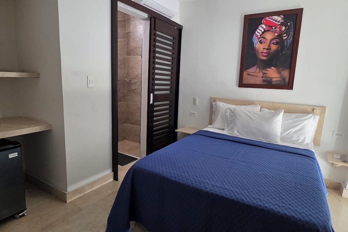 Quarto com cama, uma pequena escrivaninha, guarda-roupa e um quadro na parede.