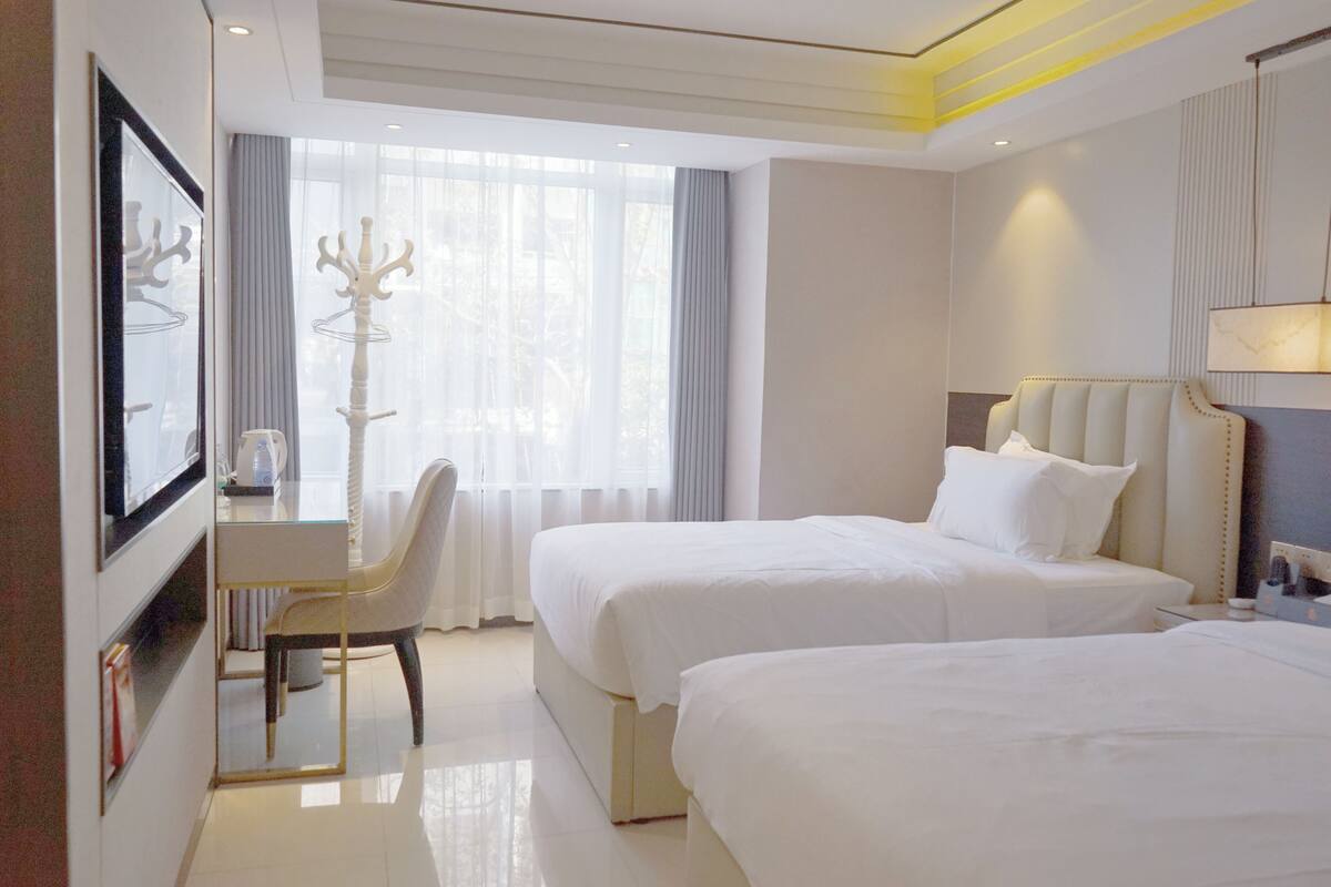 Quarto de hotel com duas camas, uma escrivaninha, uma cadeira e um cabideiro.