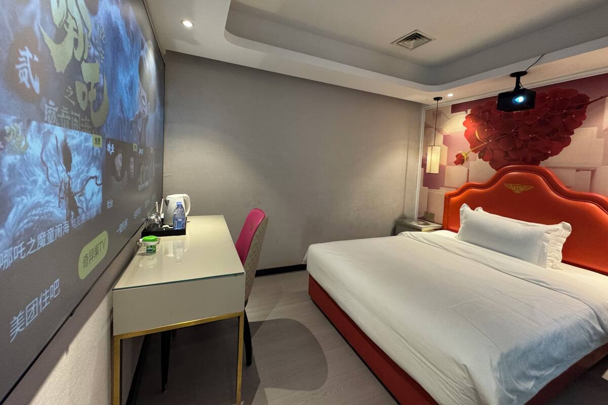 Quarto de hotel com cama, escrivaninha, cadeira, televisão e projetor.