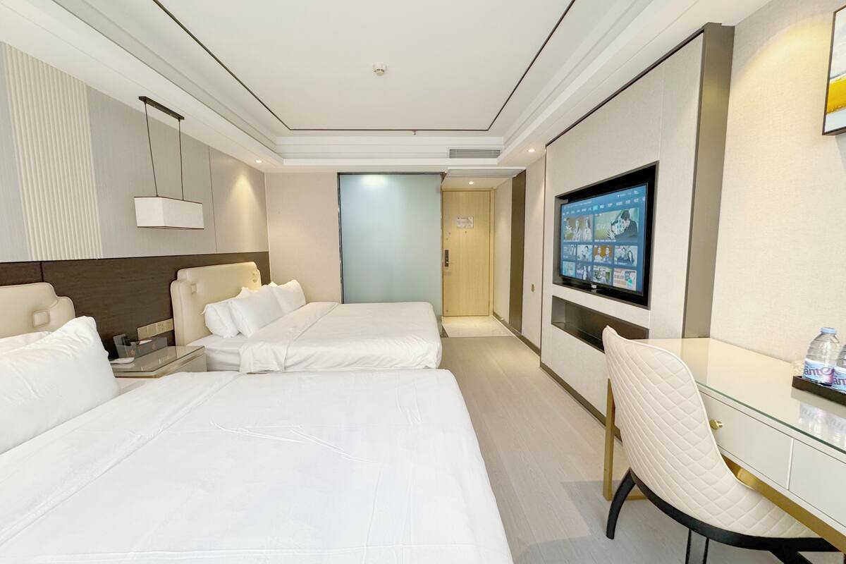 Quarto de hotel com duas camas, uma escrivaninha, uma cadeira, uma televisão e uma porta que dá para um corredor.