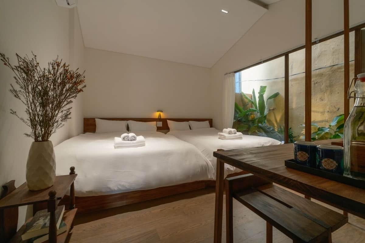 Quarto de hotel com duas camas, uma mesa de madeira, um vaso com galhos e uma janela grande com vista para vegetação.