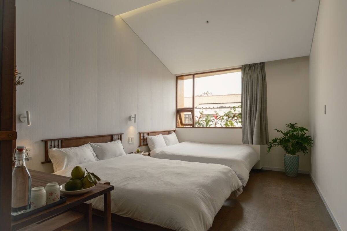 Quarto de hotel com duas camas, uma janela grande com vista, mesa de cabeceira com uma garrafa e frutas, e uma planta em vaso.