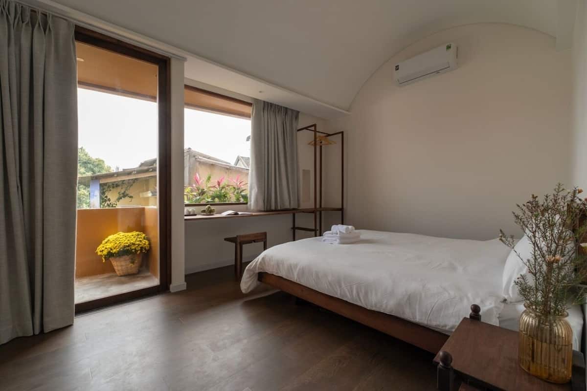 Um quarto com cama, janela com cortinas, mesa de cabeceira e um vaso com planta.