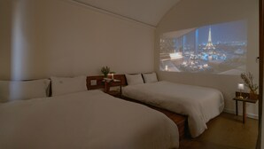 Quarto de hotel com duas camas, uma mesinha de cabeceira, um abajur e uma janela grande exibindo uma projeção da Torre Eiffel.