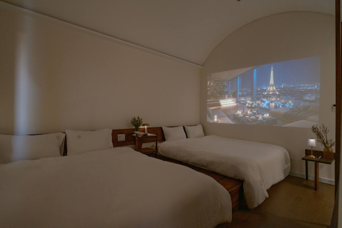 Quarto de hotel com duas camas, uma mesinha de cabeceira, um abajur e uma janela grande exibindo uma projeção da Torre Eiffel.