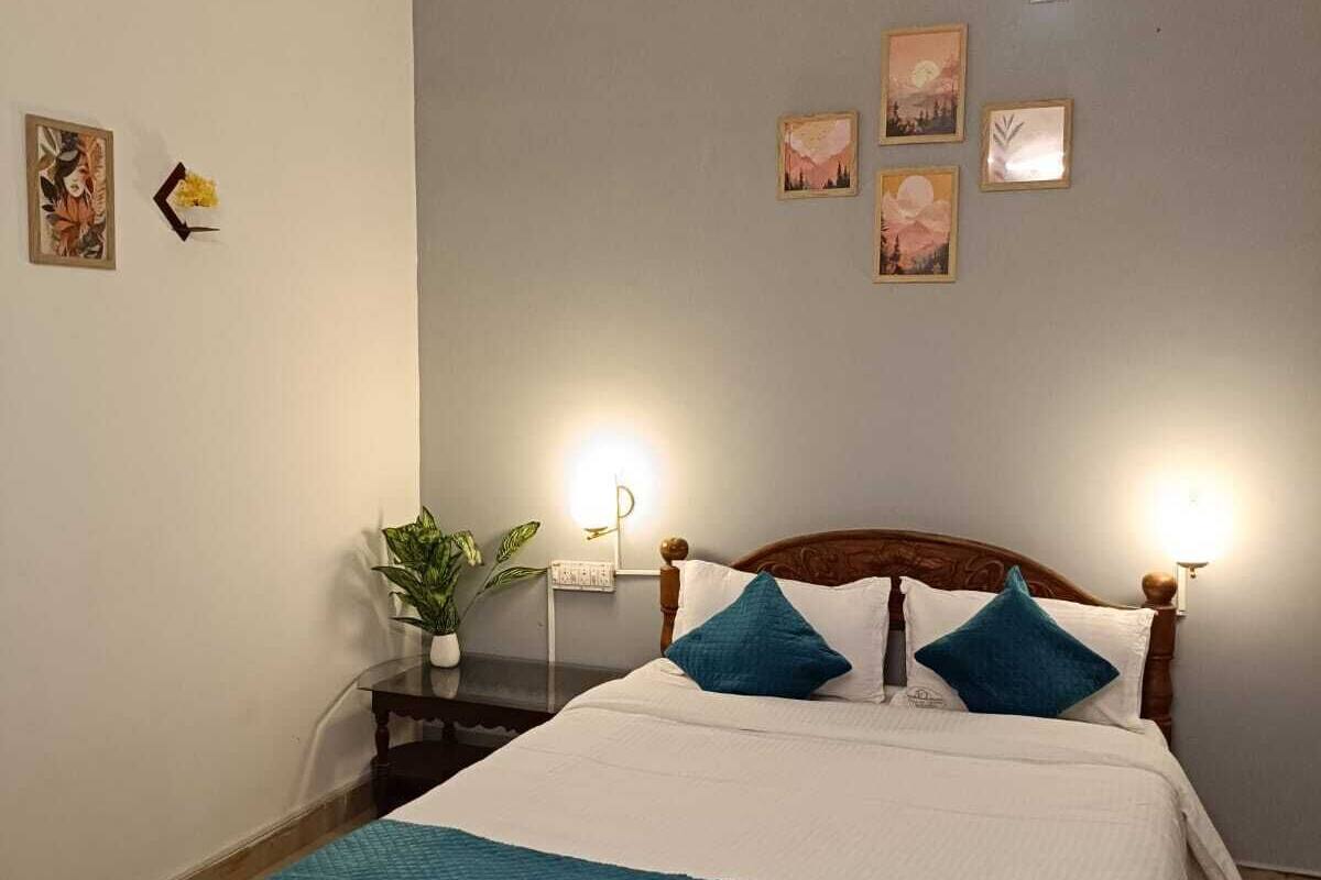 Cama bem arrumada com estrutura de madeira e roupa de cama branca, uma mesinha de cabeceira com abajur e um ventilador de teto.