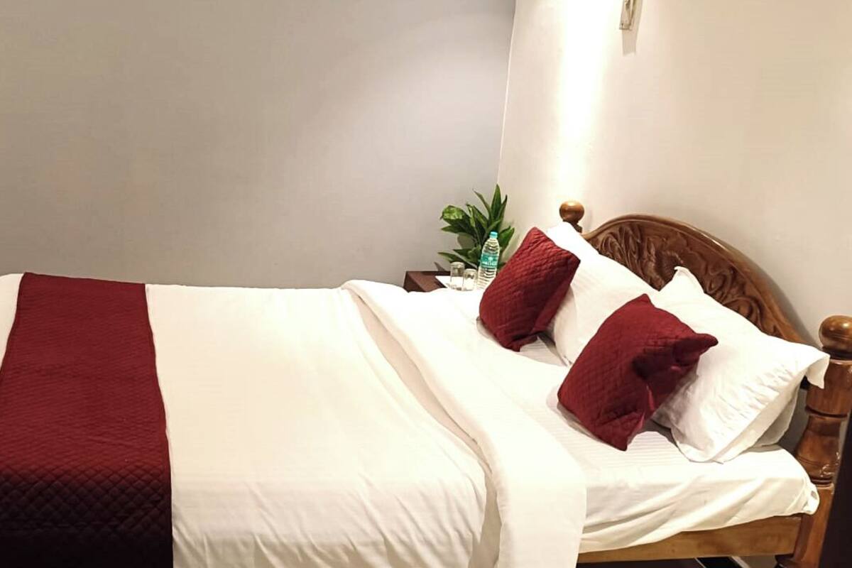 Uma cama bem arrumada com roupa de cama branca e vermelha, uma cabeceira de madeira e uma mesinha de cabeceira com uma garrafa de água e uma planta.