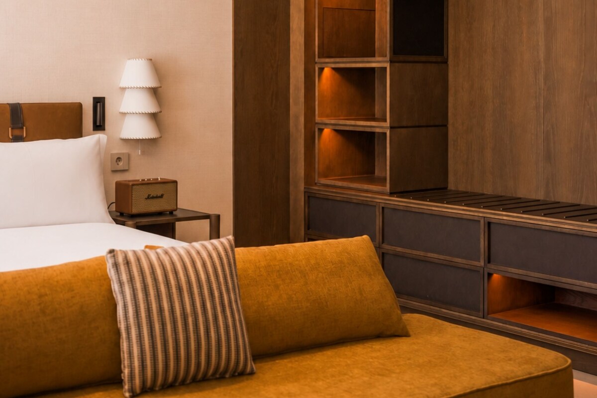 Quarto de hotel com cama, luminária de cabeceira, mesinha com rádio, sofá com almofada listrada e prateleiras embutidas.