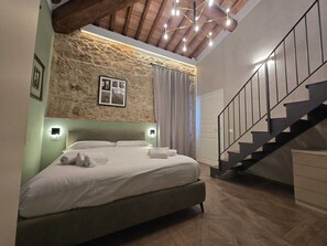 Quarto com parede de pedra, cama com roupa de cama branca, escada e luminária no teto.