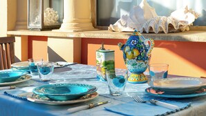 Mesa posta com pratos azuis, copos e um bule, decorada com conchas e uma janela ao fundo.