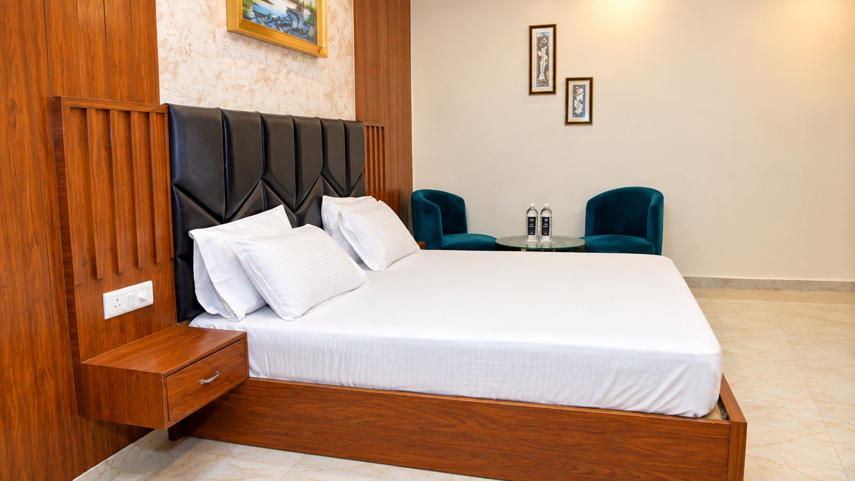 Quarto de hotel com estrutura de cama de madeira, mesa de cabeceira e duas cadeiras verde-água.