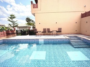 Piscina com degraus revestidos, rodeada por espreguiçadeiras e um edifício com fachada bege.