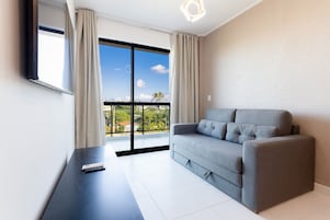 Uma sala de estar moderna com sofá cinza, televisão de tela plana na parede, uma janela grande com cortinas e uma varanda com vista para palmeiras.
