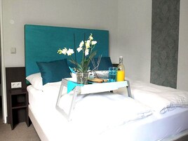 Uma cama bem arrumada com cabeceira azul-turquesa, uma mesa de cabeceira com abajur, um tabuleiro com suco de laranja, um copo de água e um vaso com flores brancas.