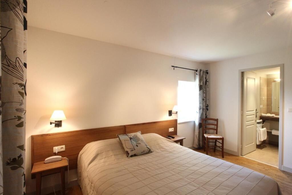 Quarto de hotel com cama, luminárias de cabeceira, uma cadeira, mesa de cabeceira e banheiro visível por uma porta aberta.