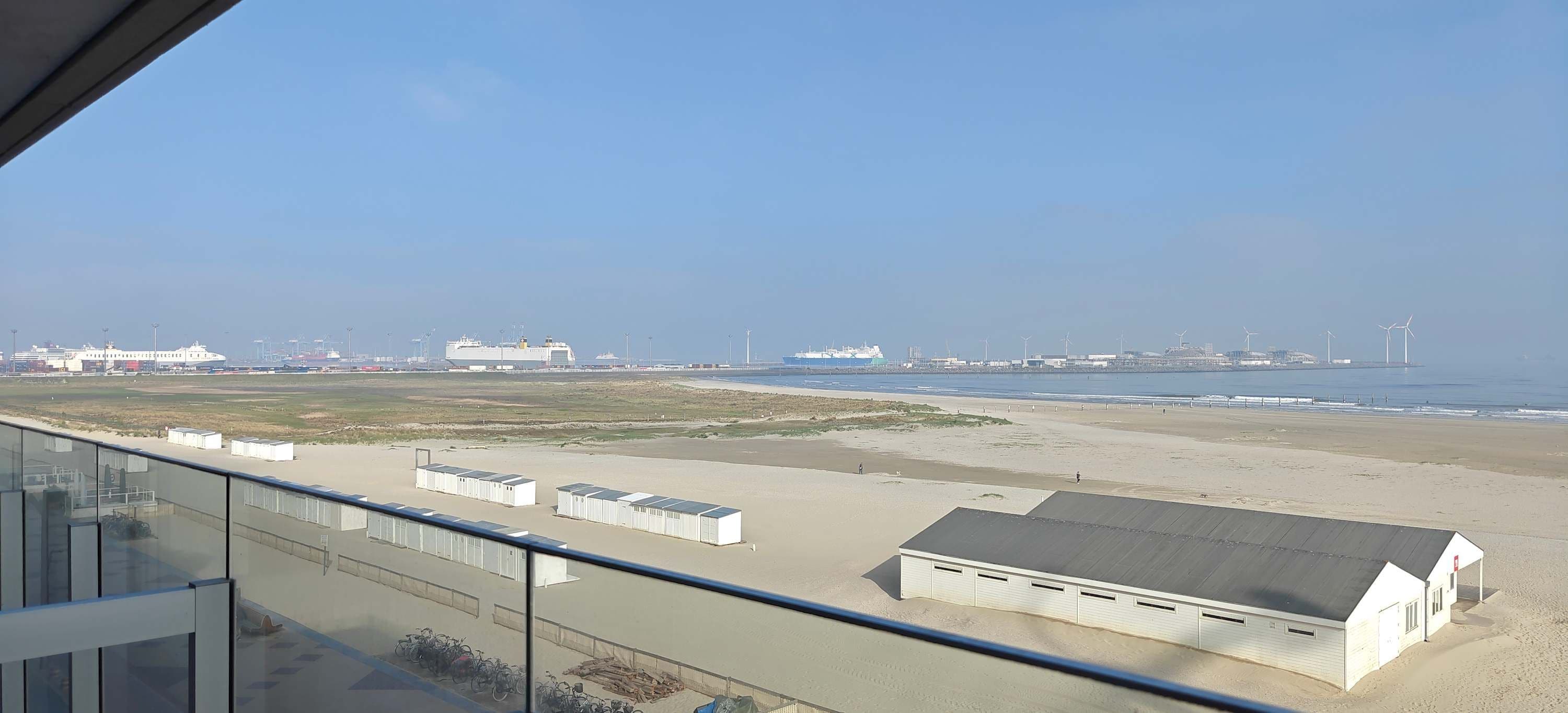 Vista de uma varanda com vista para uma praia, com alguns edifícios, incluindo uma construção branca com telhado escuro.